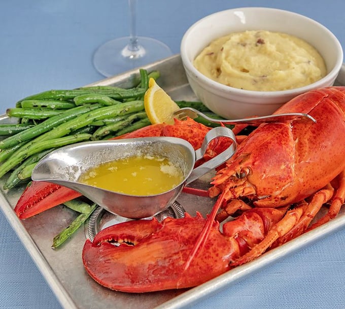A perfectly steamed lobster flanked by green beans and mashed potatoes. The silver sauce boat holds liquid gold—clarified butter for dunking.