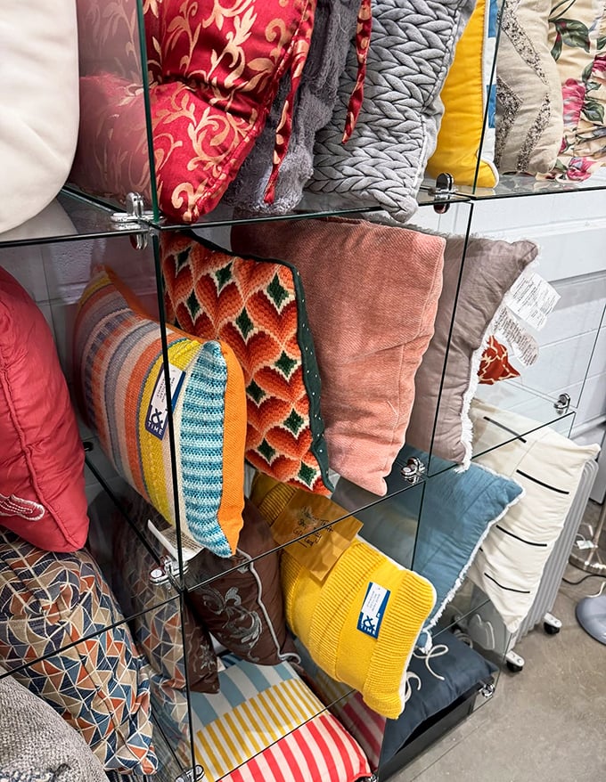 A kaleidoscope of decorative pillows stacked neatly behind glass – from vintage patterns to modern designs, each one waiting for its second chance to shine in someone's home.