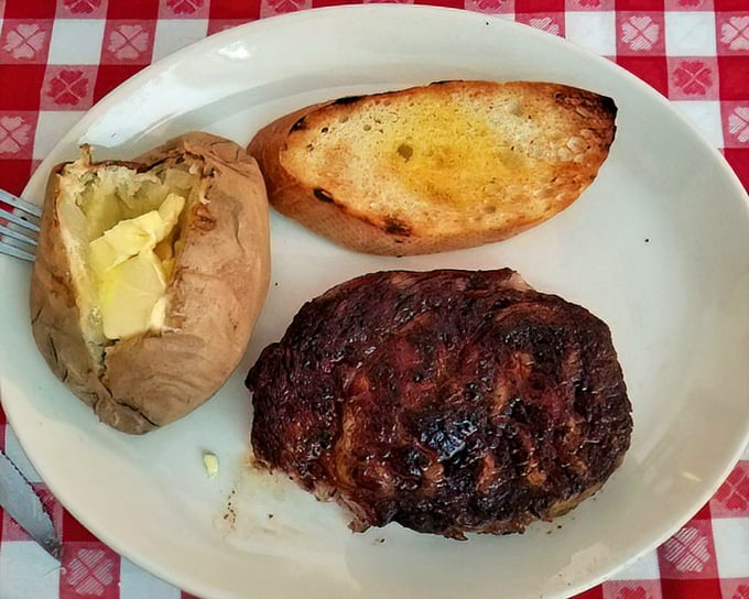 The holy trinity of steakhouse perfection: a crusty, perfectly-cooked ribeye, a butter-drenched baked potato, and garlic bread that would make an Italian grandmother nod in approval.