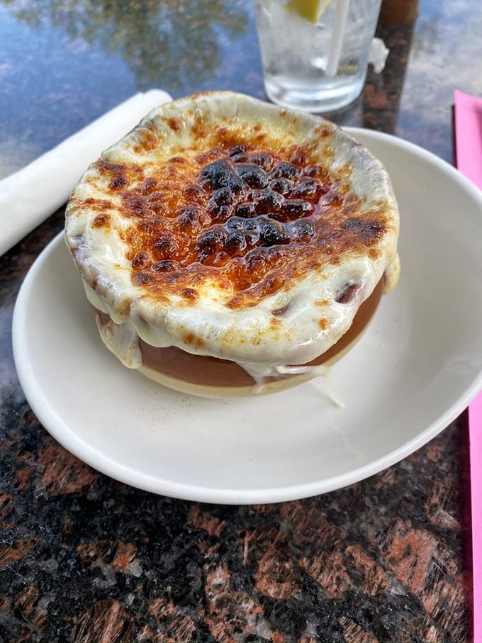 The French onion soup arrives like a cheese volcano that erupted in heaven – that perfect golden dome hiding a savory treasure worth every stretchy, messy bite.