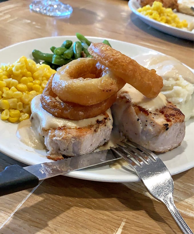 The pork chop arrives like a work of art—topped with crispy onion rings that serve as both garnish and the perfect excuse for finger food.