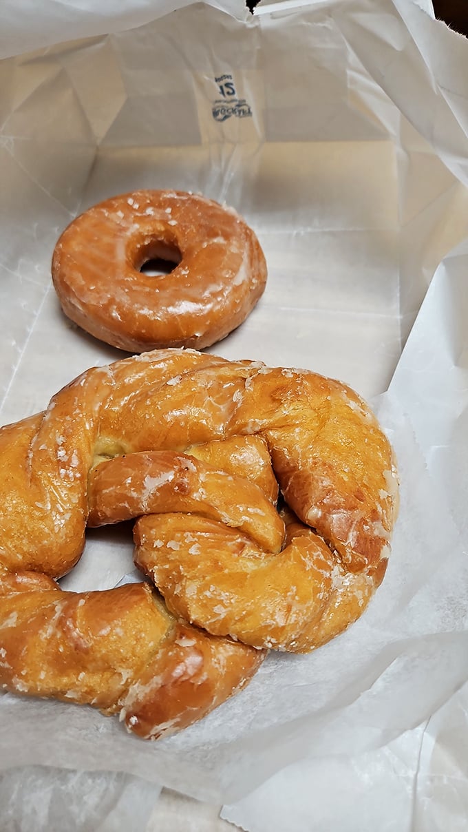 Behold the twisted geometry of perfection—a glazed pretzel donut and its circular companion, resting on wax paper like artifacts from the Museum of Deliciousness.
