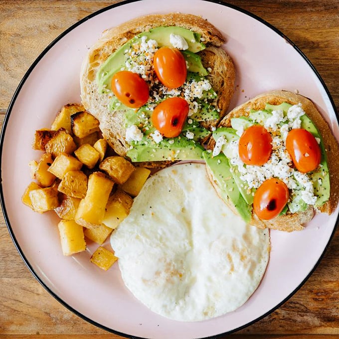 Avocado toast that Instagram was invented for—creamy avocado, bright tomatoes, and crumbled cheese on perfectly toasted bread with golden potatoes on the side.