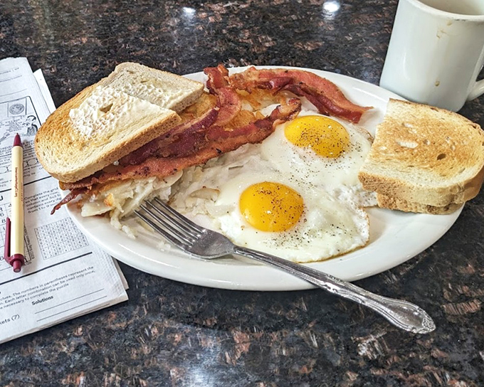 The breakfast trinity: perfectly fried eggs, crispy bacon, and hash browns that crackle with each forkful—morning salvation on a white plate.