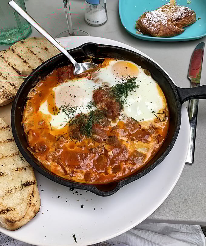Shakshuka served in a sizzling skillet—eggs nestled in spiced tomato sauce with grilled bread standing by, ready for its supporting role in this Mediterranean masterpiece.