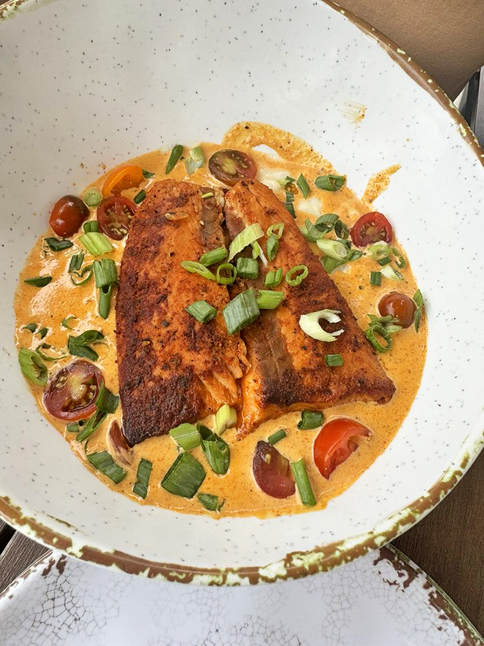 Blackened salmon lounging in a creamy pool of sauce, garnished with green onions and cherry tomatoes—like a spa day for your taste buds.