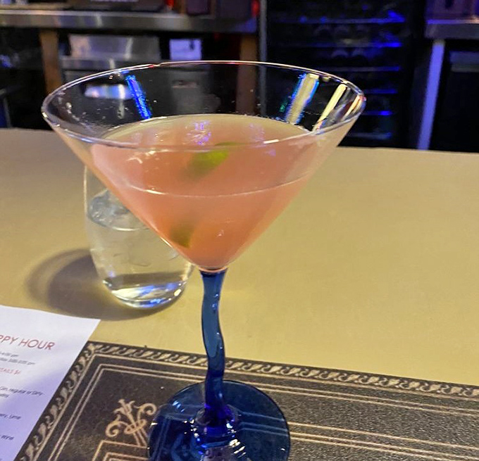 The cocktail glows with possibility in its blue-stemmed glass—like a sunset captured in liquid form, promising good decisions or interesting stories.